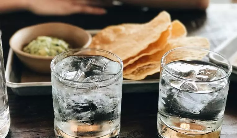 Dois copos de água em uma mesa, com comida ao fundo.