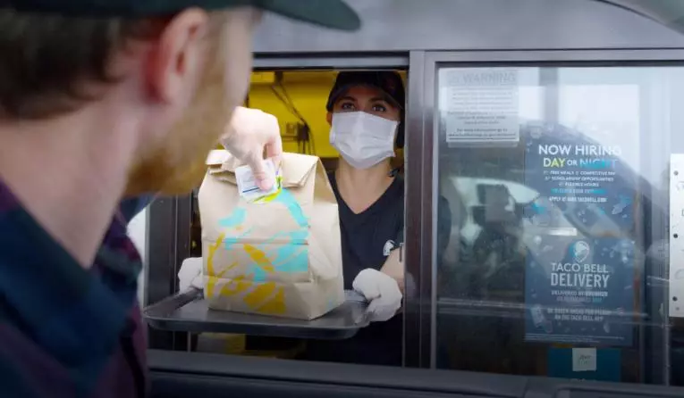 „Taco Bell“ važiuoja pro kaukę dėvintį darbuotoją, kuris klientui perduoda maistą.