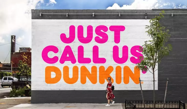 En kvinde går forbi et vægmaleri, hvor der står Just Call Us Dunkin'.